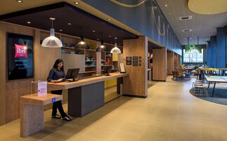 Uma área de recepção de hotel com tela digital exibindo informações do hotel, balcão de atendimento e área de estar com mesas e cadeiras.