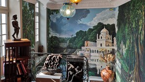 Quarto com um mural de paisagem, uma estátua sobre uma mesa e uma estante de livros.