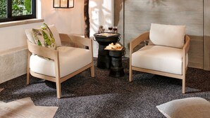 Quarto com duas cadeiras de madeira, uma mesa pequena com uma vela, um letreiro que diz 'nosso jardim' e uma janela grande com vegetação lá fora.
