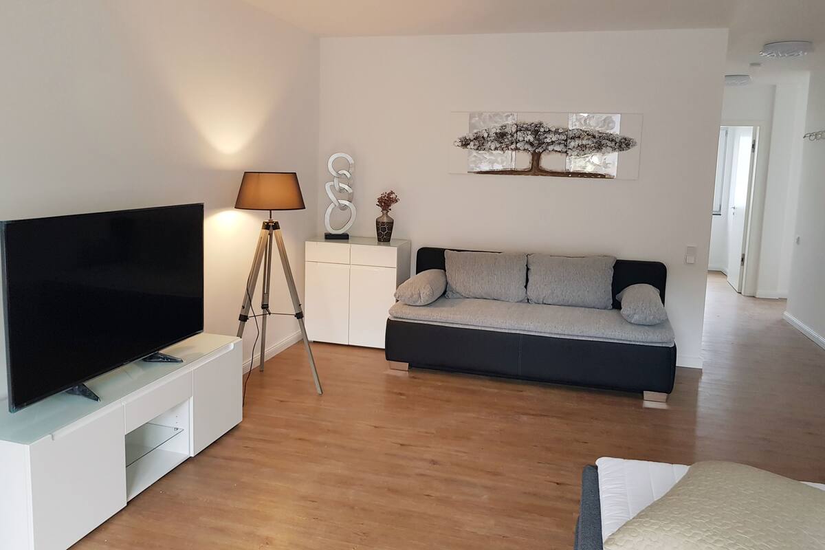 Uma sala de estar moderna com televisão de tela plana, luminária de tripé, armário branco, sofá cinza com almofadas e um objeto decorativo na parede.