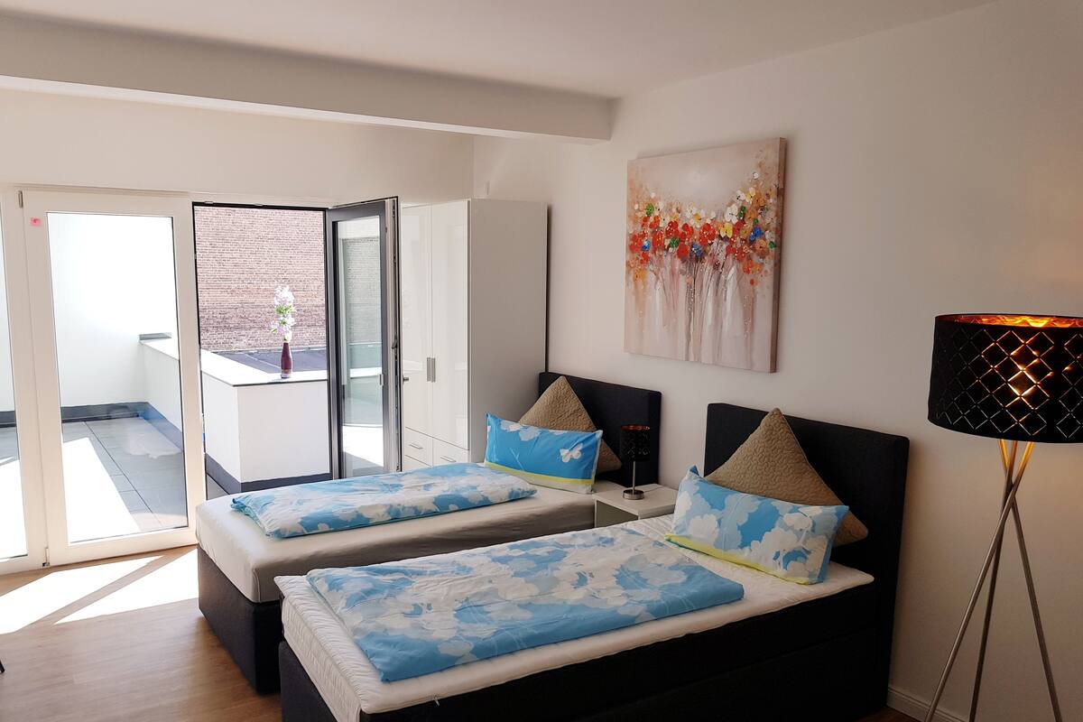 Quarto moderno com duas camas, porta de vidro deslizante para uma varanda, um quadro na parede e um abajur.