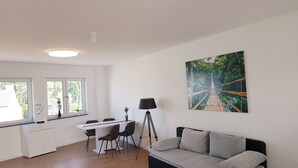 Uma sala de estar moderna com sofá, mesa de jantar com cadeiras, abajur e uma grande pintura de uma ponte em uma floresta.