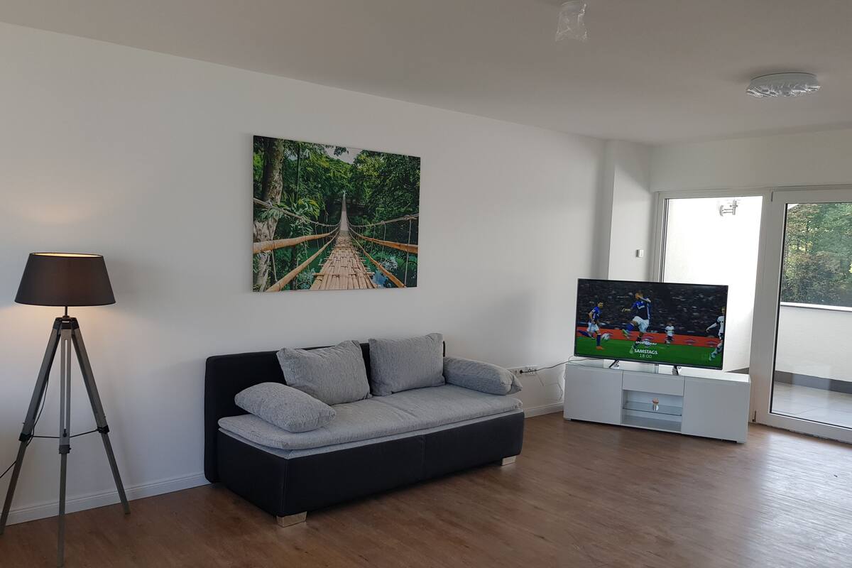 Uma sala de estar com um sofá preto, um abajur, uma televisão sobre um suporte branco e uma grande pintura de uma ponte.