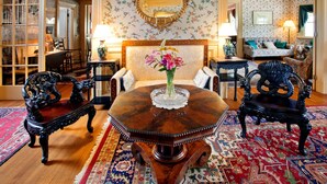 Quarto tradicional com mesa de madeira redonda, cadeiras ornamentadas, papel de parede floral e um espelho grande na parede.