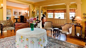 Um quarto com uma mesa redonda coberta por um tecido floral, cercada por cadeiras estampadas, um tapete grande e padronizado, e um vaso com flores.