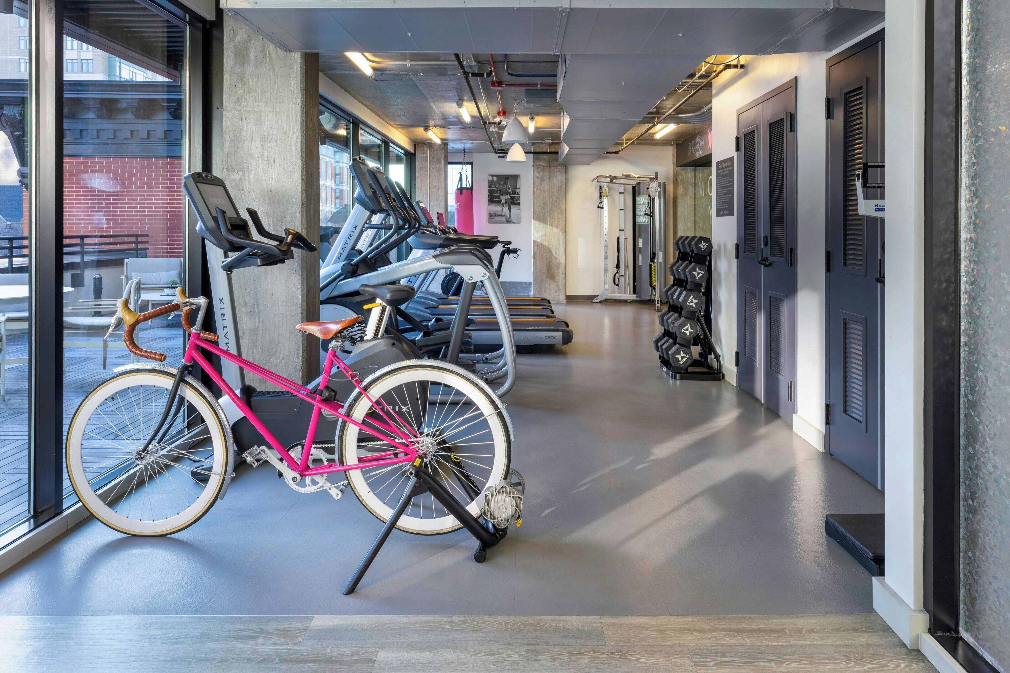 Uma academia com equipamentos de exercício, uma bicicleta rosa e vista para o exterior.