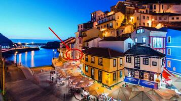 Uma vila costeira ao anoitecer, com calçadão à beira-mar, edifícios com varandas e áreas de estar ao ar livre.