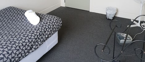 Um quarto pequeno e bem organizado, com uma cama, uma mesa de cabeceira, um cabideiro, um ventilador e uma porta com um aviso pendurado.