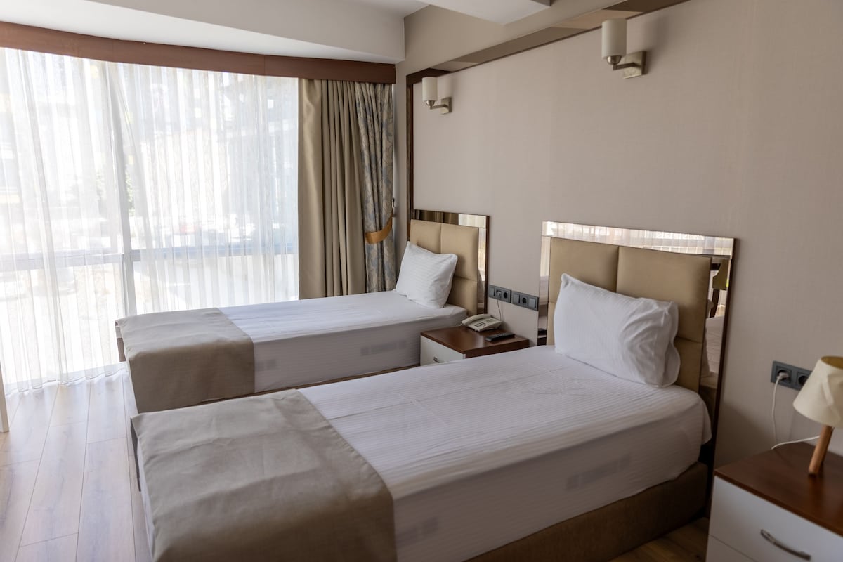 Quarto de hotel com duas camas, uma mesa de cabeceira com telefone e uma janela com cortinas.