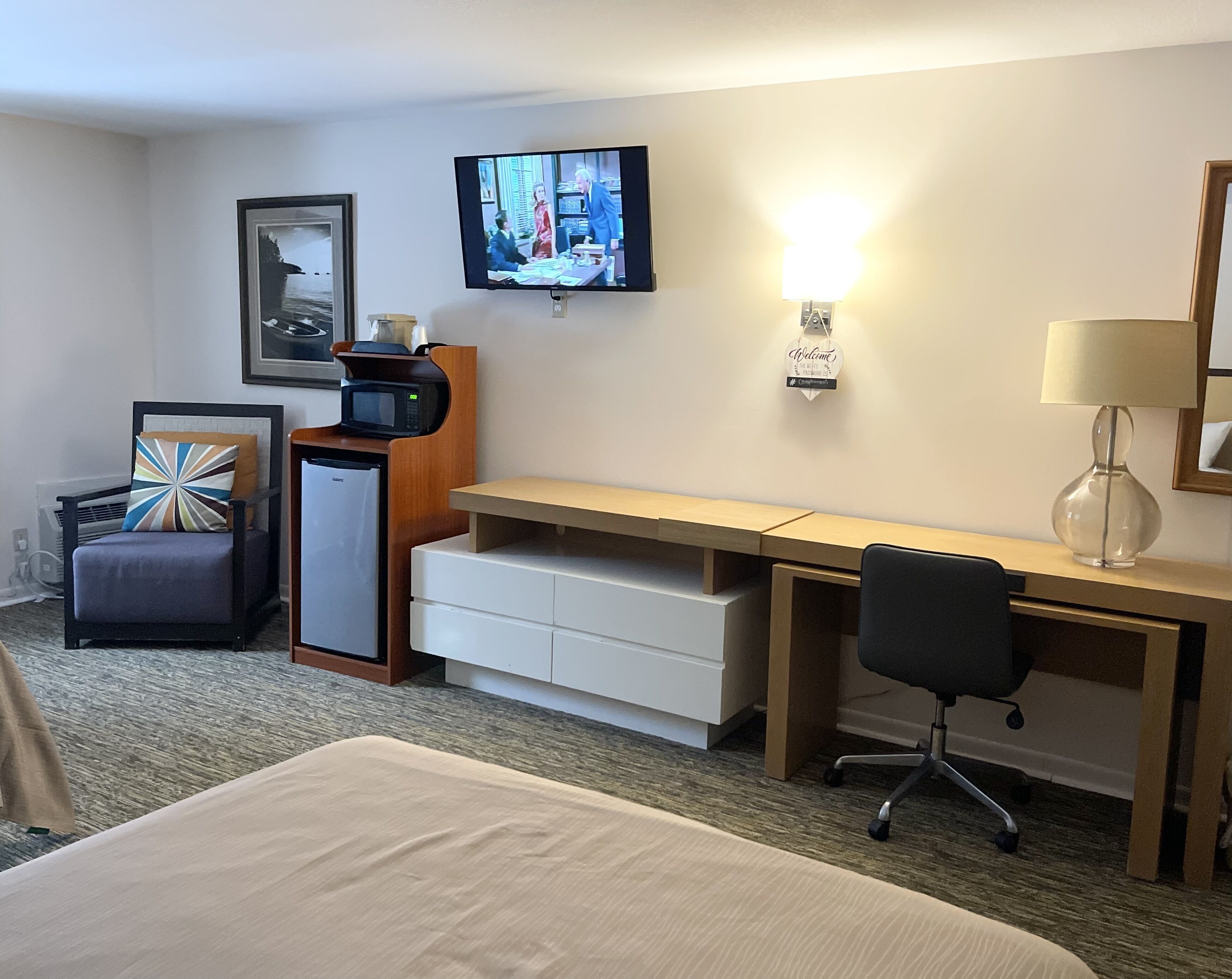 Quarto de hotel com uma cama, escrivaninha com gavetas, uma cadeira, televisão fixada na parede, quadro decorativo e placa de boas-vindas.