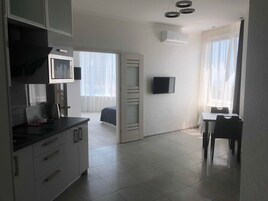 Quarto de hotel moderno com cozinha compacta, televisão de tela plana na parede, área de refeições com mesa e cadeiras, e uma grande janela com cortinas translúcidas.