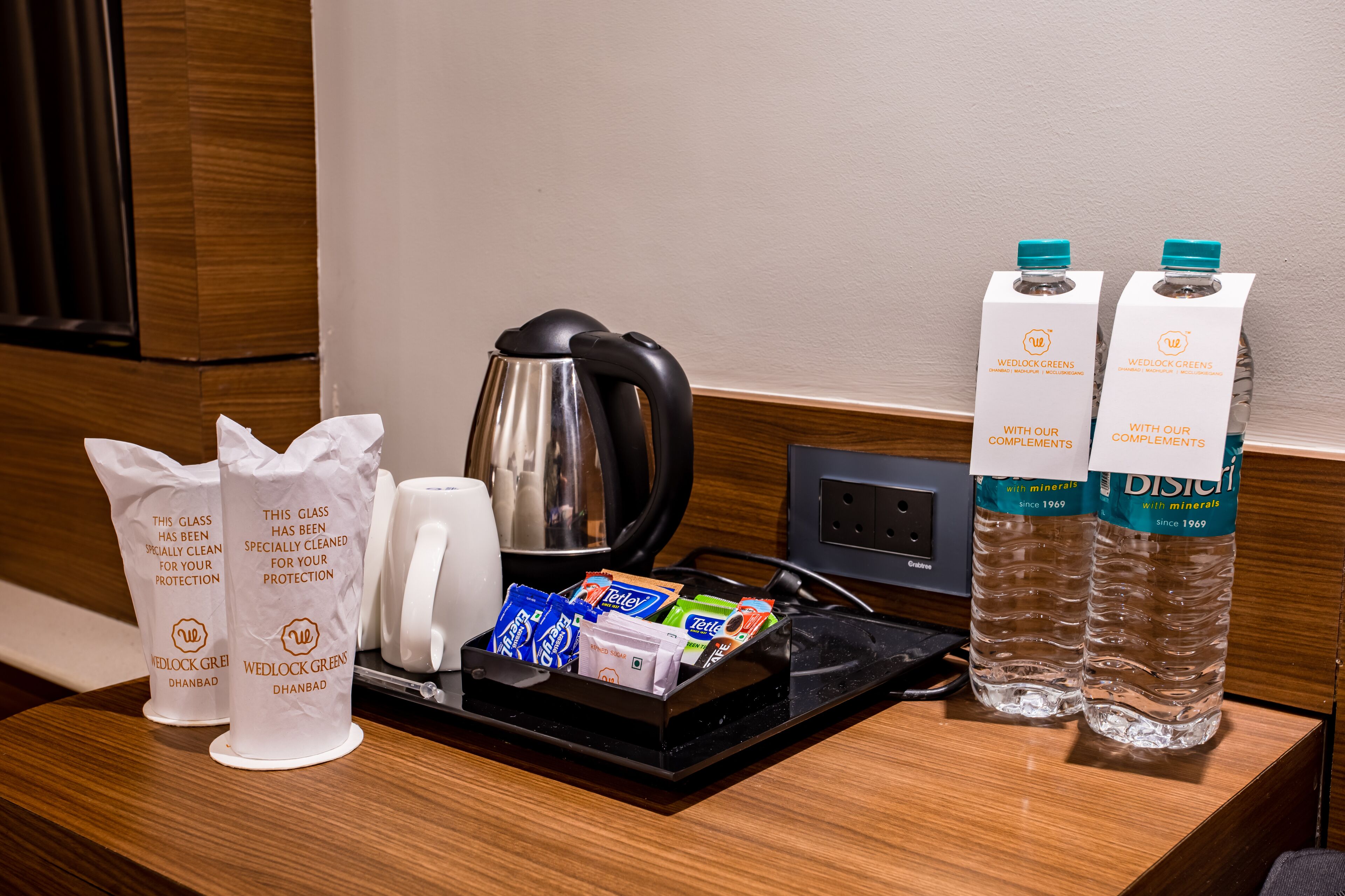 Uma área de recepção de hotel com um canto para café, incluindo uma chaleira, sachês de café e uma bandeja. Há duas garrafas de água e dois recipientes de vidro com a inscrição 'Wedlock Greens' no balcão.
