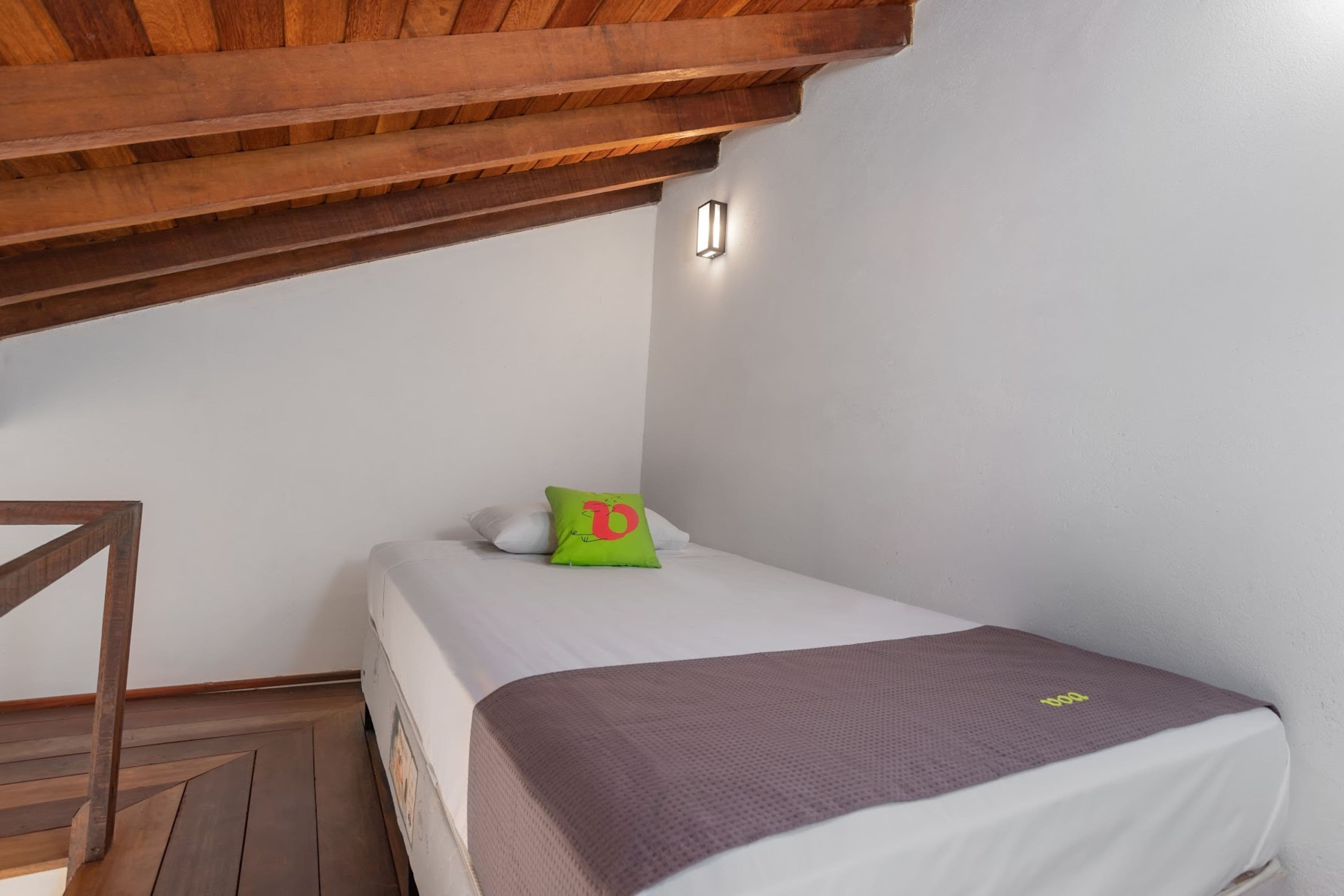 Uma cama pequena e bem arrumada com colchão branco e manta roxa, um travesseiro verde com uma letra vermelha, e um aplique de luz na parede.