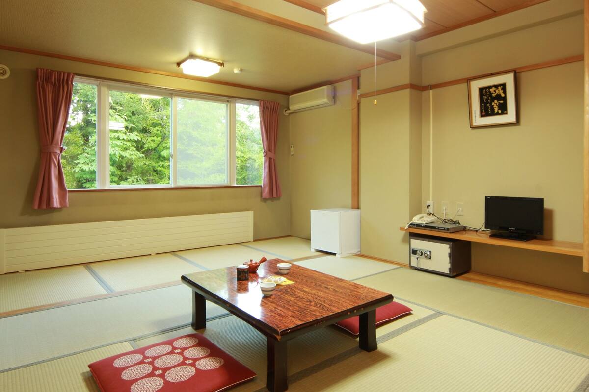 Um quarto japonês tradicional com tatames, uma mesa baixa com almofadas, uma televisão e uma janela com cortinas.