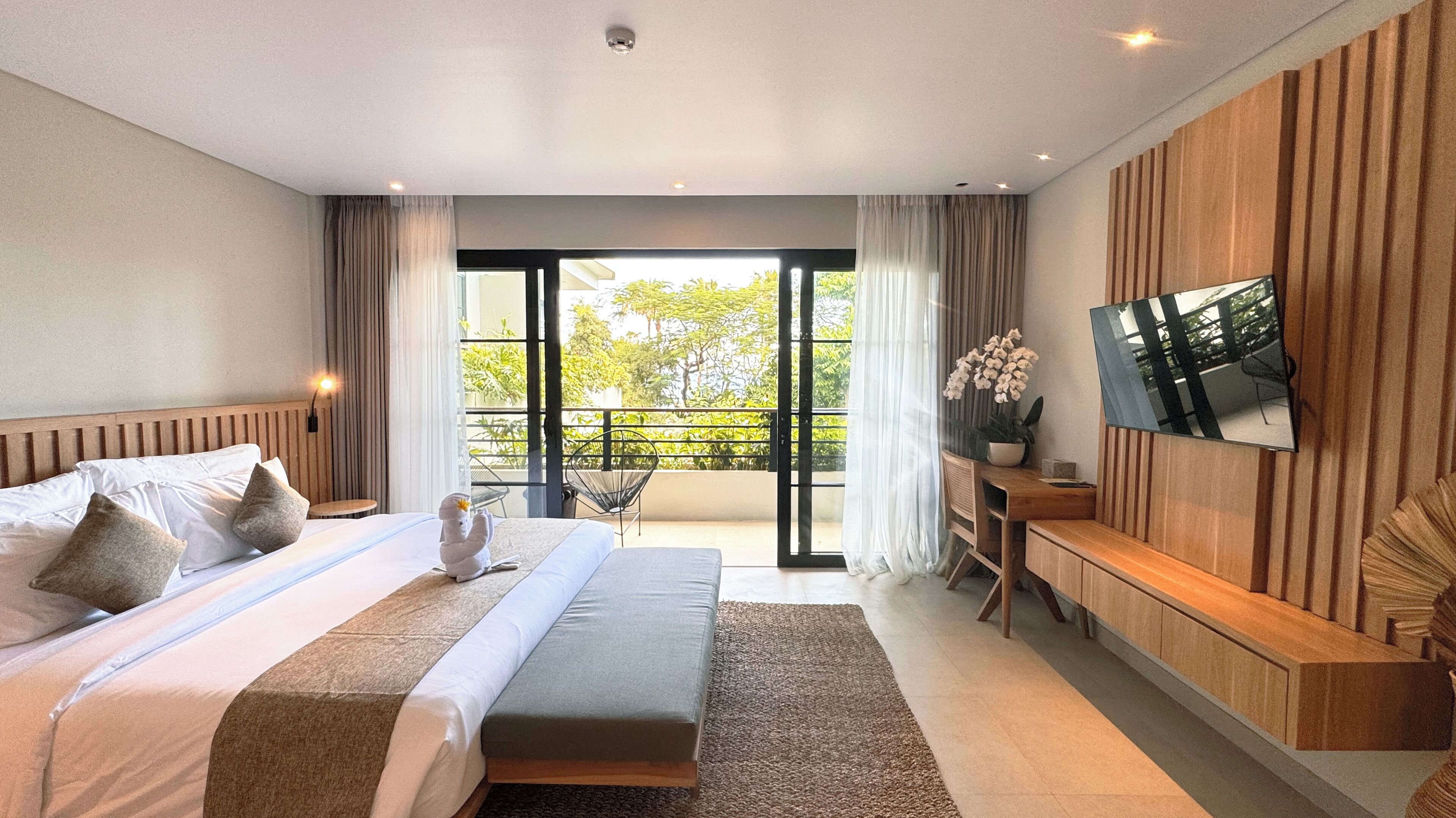 Quarto de hotel moderno com uma cama grande, uma TV na parede, varanda com vista para o verde e uma pequena escrivaninha com cadeira.