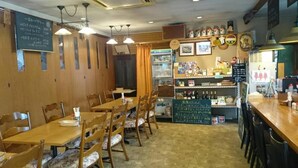 Interior de restaurante com mesas e cadeiras de madeira, um balcão com menu e prateleiras com garrafas e decorações.