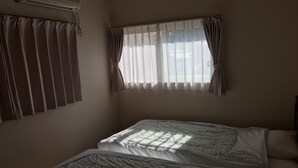 Um quarto pequeno e com pouca luz, com duas camas, uma janela com cortinas leves e um aparelho de ar condicionado na parede.