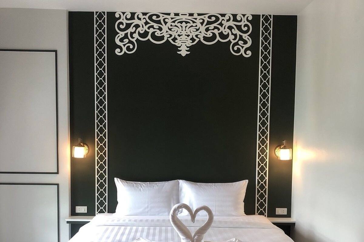 Cama bem arrumada com toalha em formato de coração nos travesseiros, cabeceira estampada em preto e branco e duas luminárias fixadas na parede.