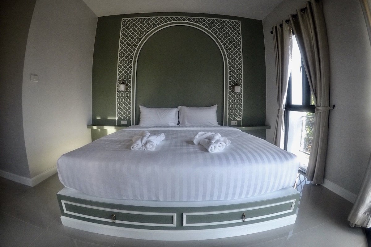 Uma cama bem arrumada com lençóis e toalhas brancas, uma janela grande com cortinas e uma parede verde com um arco decorativo.