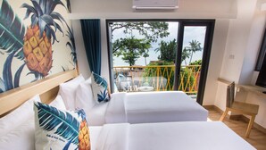 Quarto de hotel com varanda, cama com roupa de cama branca, travesseiro com tema tropical e vista para o mar e palmeiras.