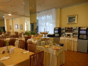 Uma área de refeições com mesas preparadas para o café da manhã, um buffet com diversas opções de alimentos e uma máquina de café.