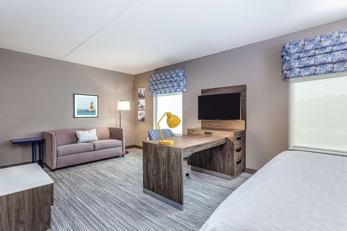 Quarto de hotel com sofá, escrivaninha com luminária, televisão e cama.