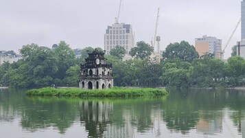 Uma estrutura tradicional em estilo de pagode, rodeada por vegetação exuberante e água, com edifícios modernos ao fundo.