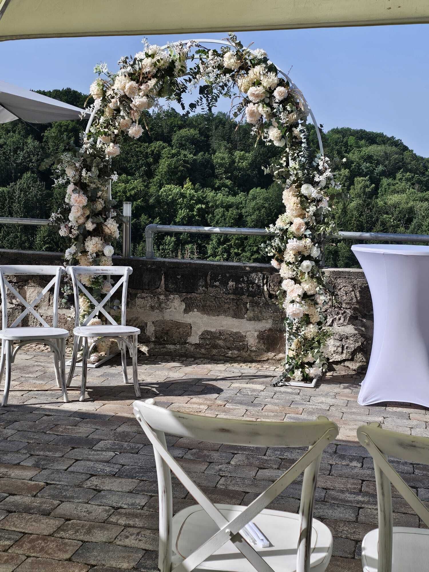 Uma área de estar ao ar livre com cadeiras brancas e um arco floral.