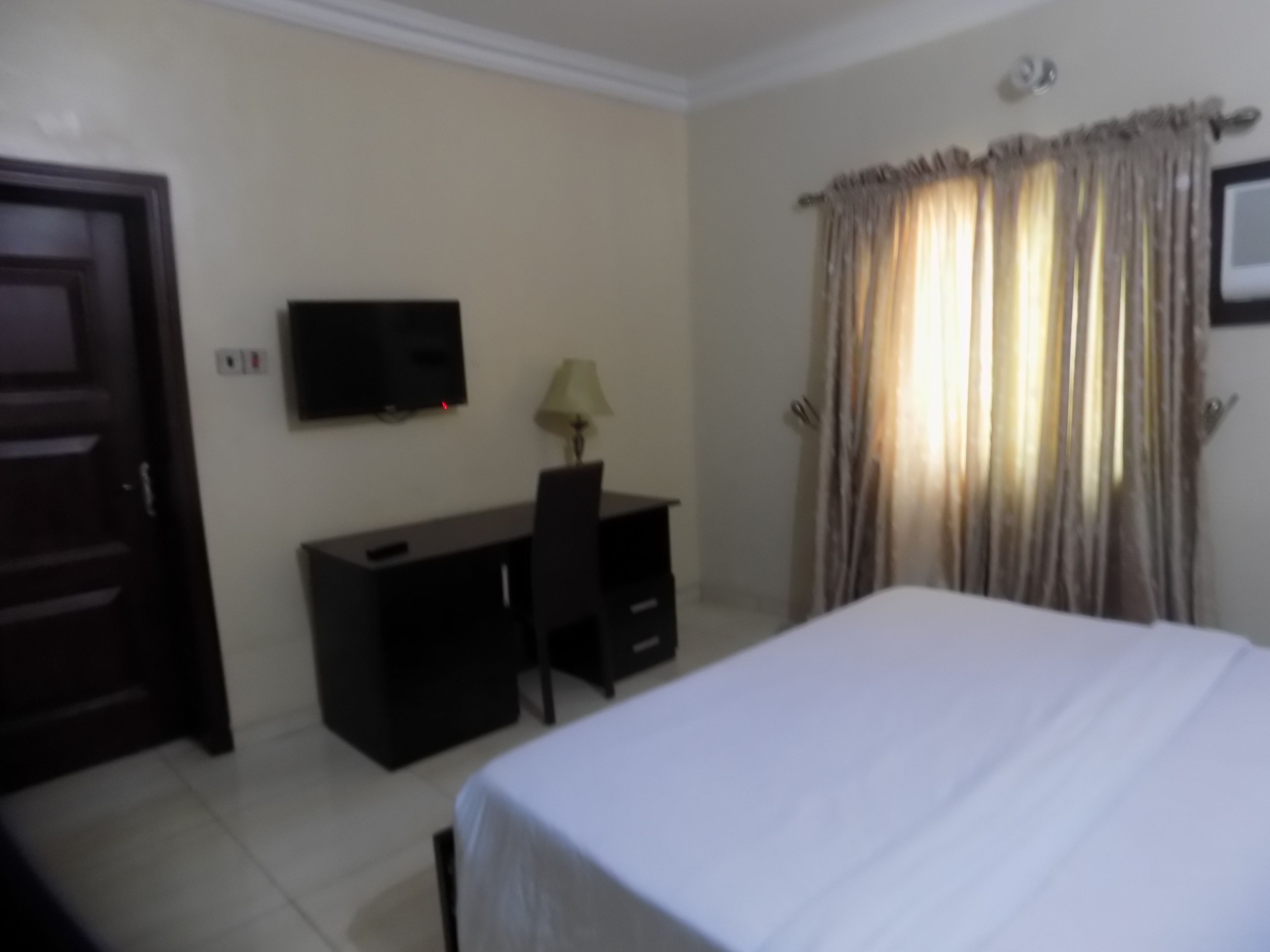 Quarto de hotel com uma cama, uma escrivaninha com cadeira, uma televisão na parede e cortinas na janela.