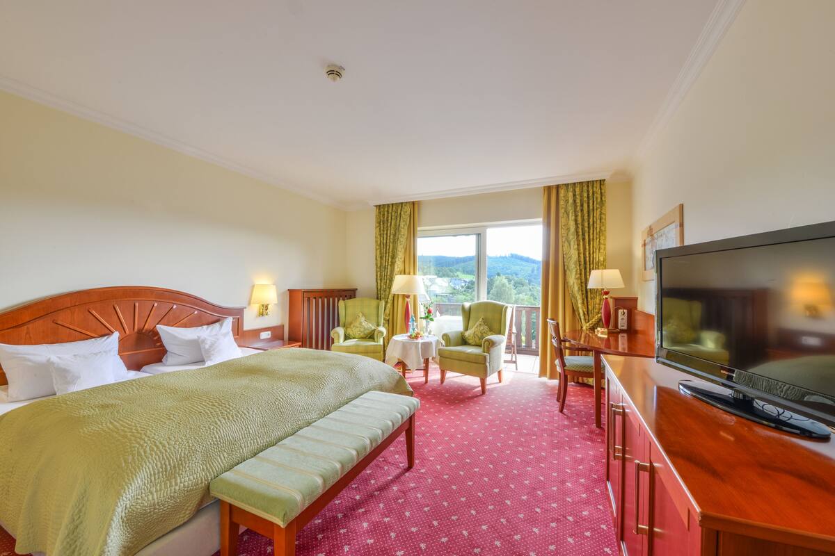 Quarto de hotel com uma cama grande, televisão, escrivaninha com cadeira e varanda com vista.
