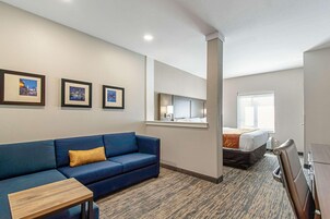 Quarto de hotel com um sofá azul, uma pequena mesa de madeira, uma cama com colcha branca e laranja, uma janela e quadros nas paredes.