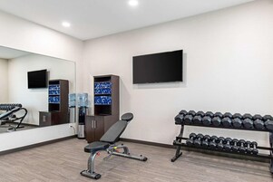 Uma academia com equipamentos de exercício, um banco, bebedouros e uma TV na parede.