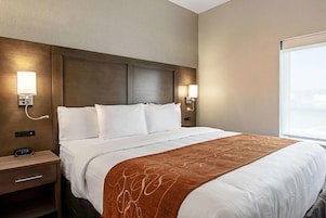 Quarto de hotel com uma cama grande, duas mesas de cabeceira, um criado-mudo, uma luminária na parede e uma janela.