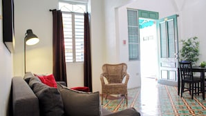 Uma sala de estar com um sofá, uma poltrona de vime, uma mesa com cadeiras, um abajur e um piso estampado.