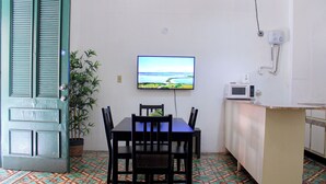 Uma área de refeições com mesa e cadeiras, uma televisão na parede, um micro-ondas na bancada e um vaso com planta.