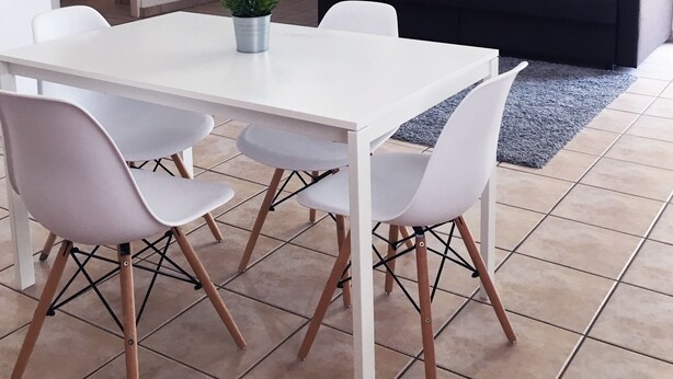 Uma sala de estar moderna com uma mesa de jantar branca e cadeiras, um sofá preto com almofada e uma pequena planta em vaso.