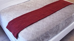 Cama bem arrumada com roupa de cama branca, manta vermelha e almofadas listradas. Há duas mesas de cabeceira com abajures, e um espelho grande na parede.