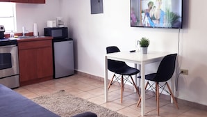 Cozinha moderna com micro-ondas, máquina de café e pequena geladeira. Há uma área de jantar com mesa branca, duas cadeiras pretas e uma pequena planta em vaso. Um ventilador de teto é visível acima da cozinha.