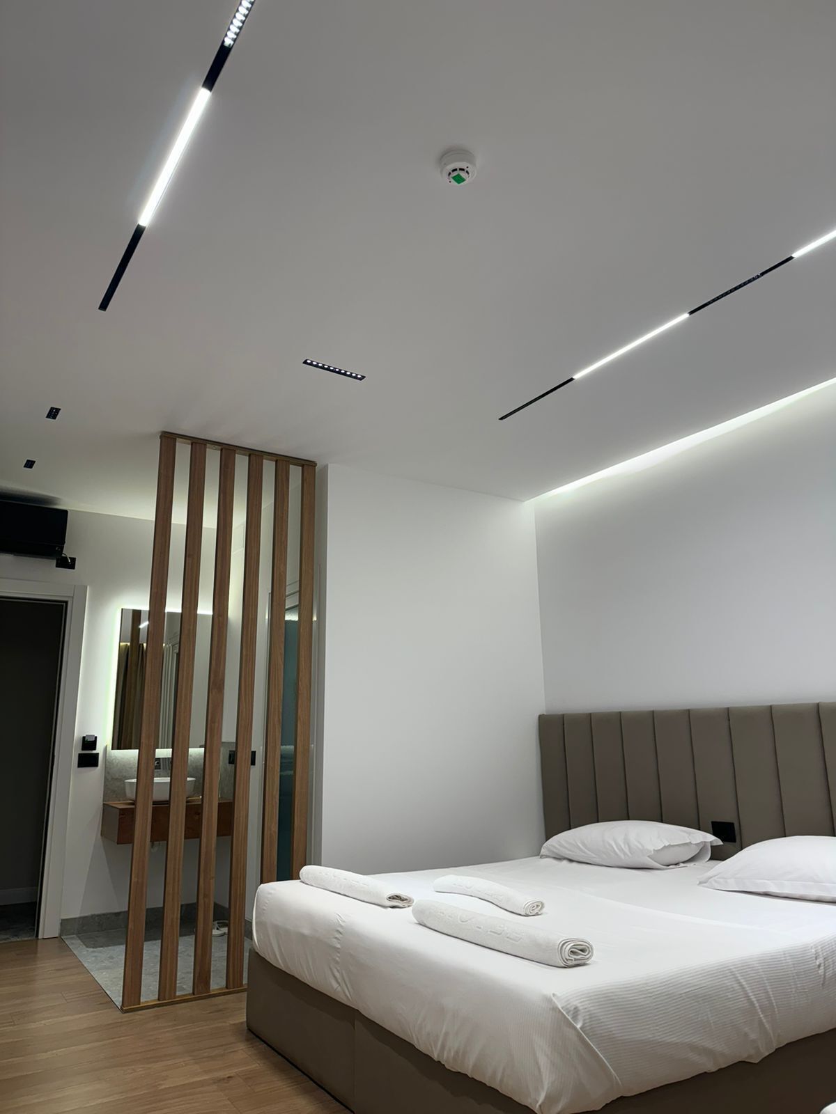 Quarto moderno com cama, divisória de madeira e teto com iluminação embutida.