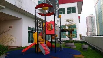 Um parquinho com escorregadores e estruturas de escalada, cercado por um edifício com janelas amplas e uma área gramada.