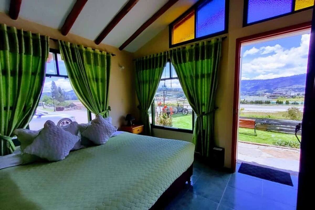 Quarto com cama, cortinas verdes e vista para uma paisagem pela janela.