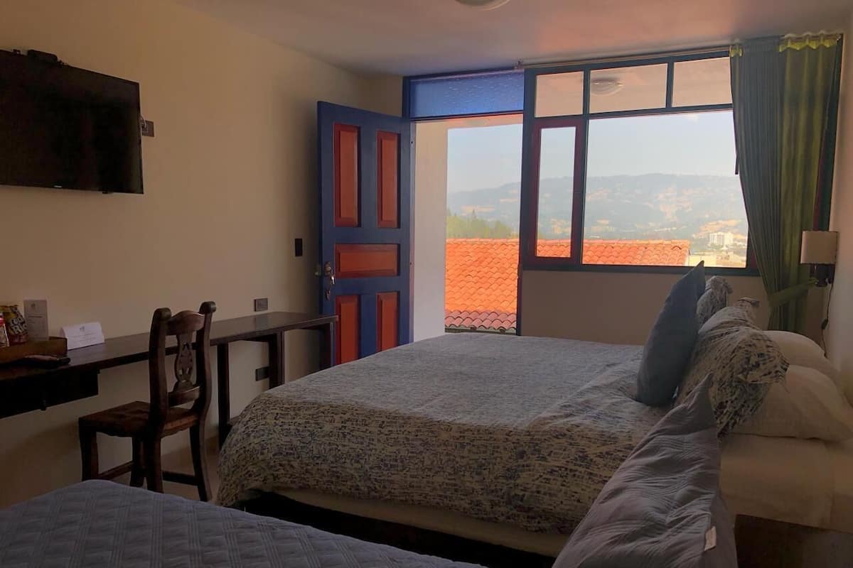 Quarto de hotel com duas camas, uma escrivaninha, uma cadeira e janela com vista para a cidade.