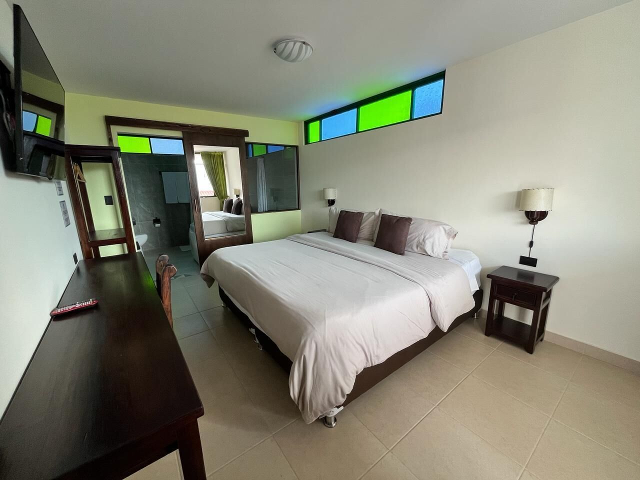 Quarto de hotel com uma cama grande, uma escrivaninha, uma cadeira, uma mesa de cabeceira e um espelho.