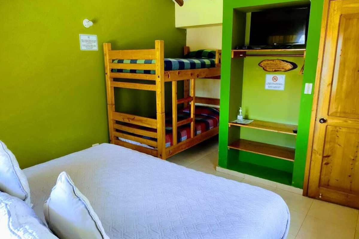 Quarto com cama beliche, uma cama de solteiro, parede verde, porta de madeira e prateleira com televisão.