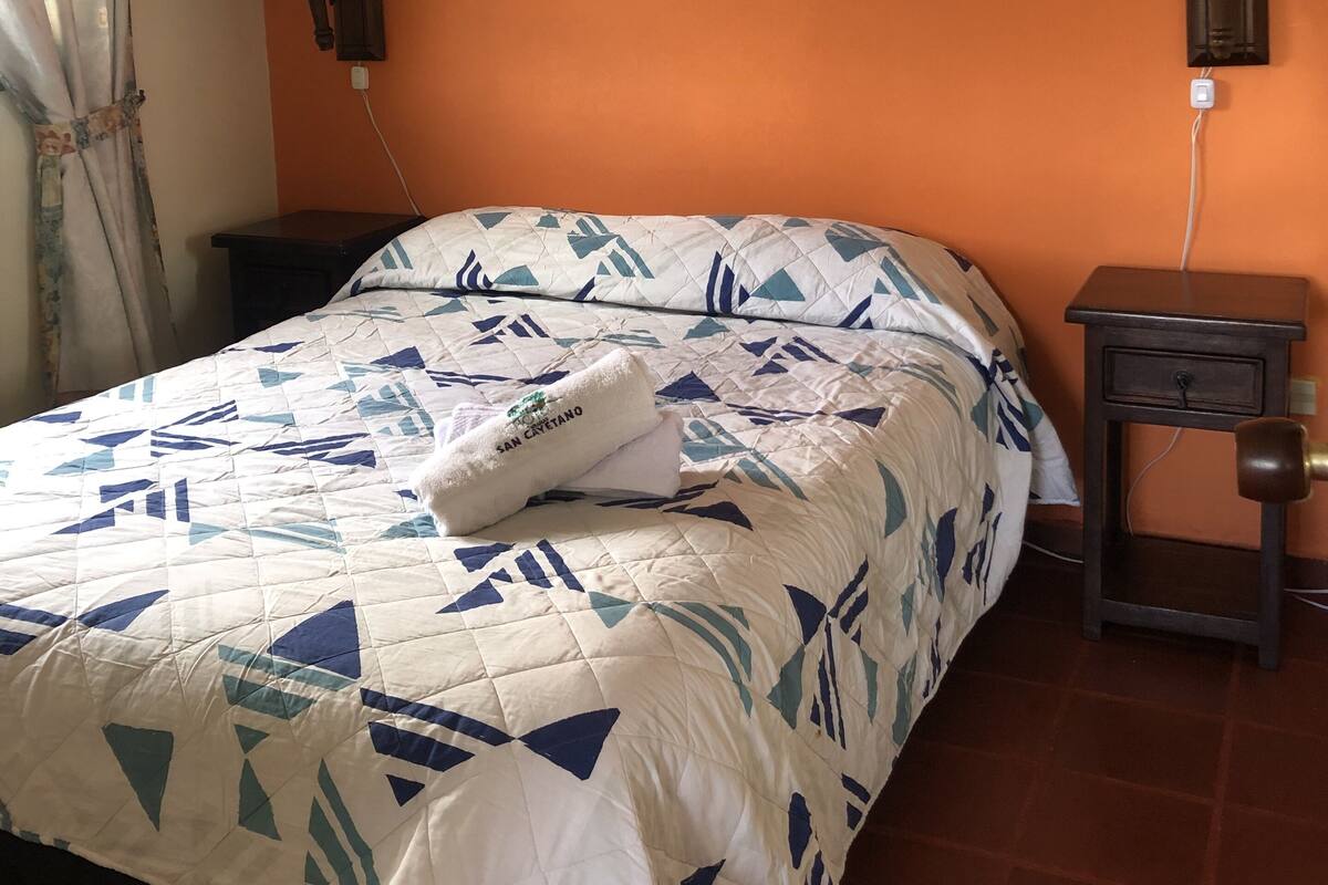 Uma cama com um edredom estampado, duas luminárias na parede, uma mesinha de cabeceira e uma cortina.