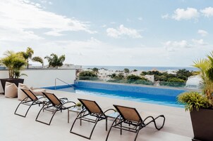 Uma área de piscina no terraço, com espreguiçadeiras, vista clara para o mar e a placa do hotel ao fundo.