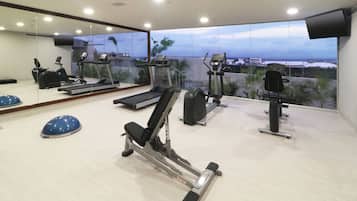 Uma academia com diversos equipamentos de exercícios, incluindo esteiras e aparelhos de musculação, e uma grande janela com vista para a paisagem urbana.