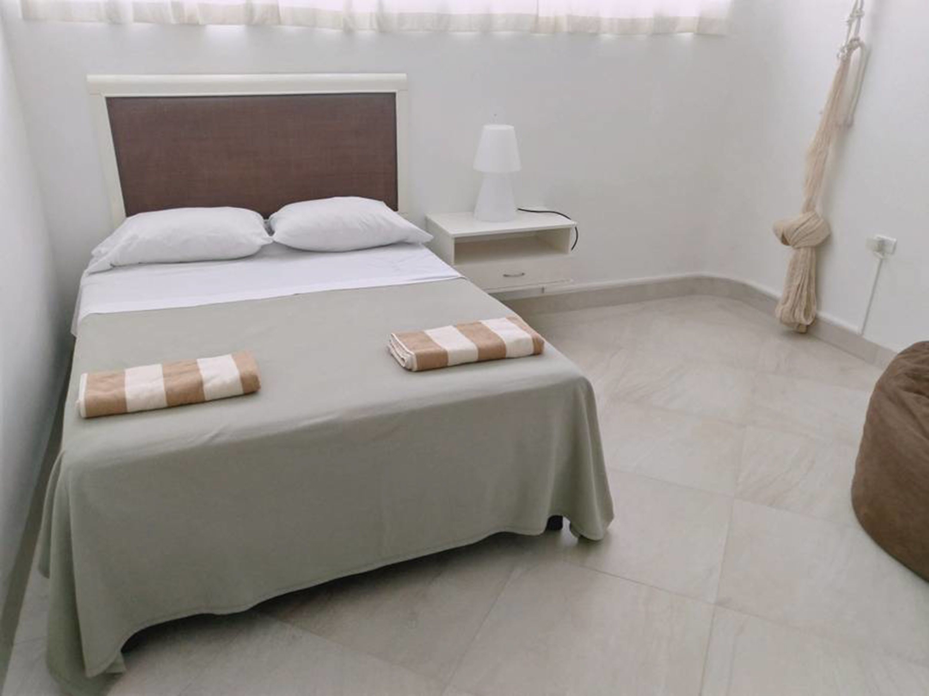Cama bem arrumada com edredom cinza e duas toalhas listradas, mesa de cabeceira com abajur e um enfeite de macramê na parede.