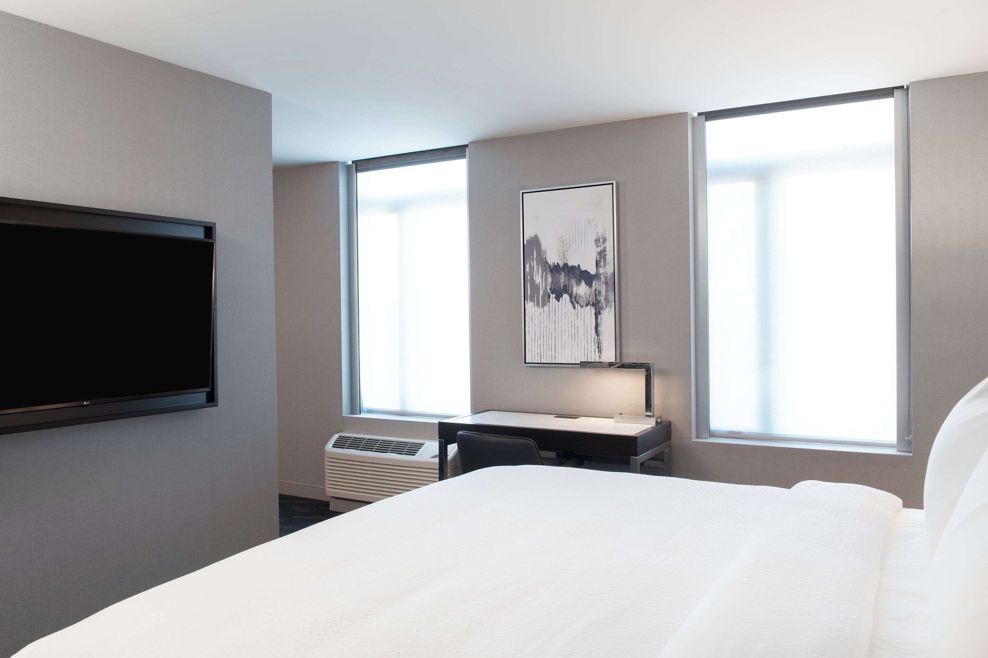 Quarto de hotel com cama, escrivaninha, televisão de tela plana, janela e obra de arte pendurada na parede.