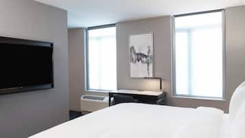Quarto de hotel com cama, escrivaninha, televisão de tela plana, janela e obra de arte pendurada na parede.